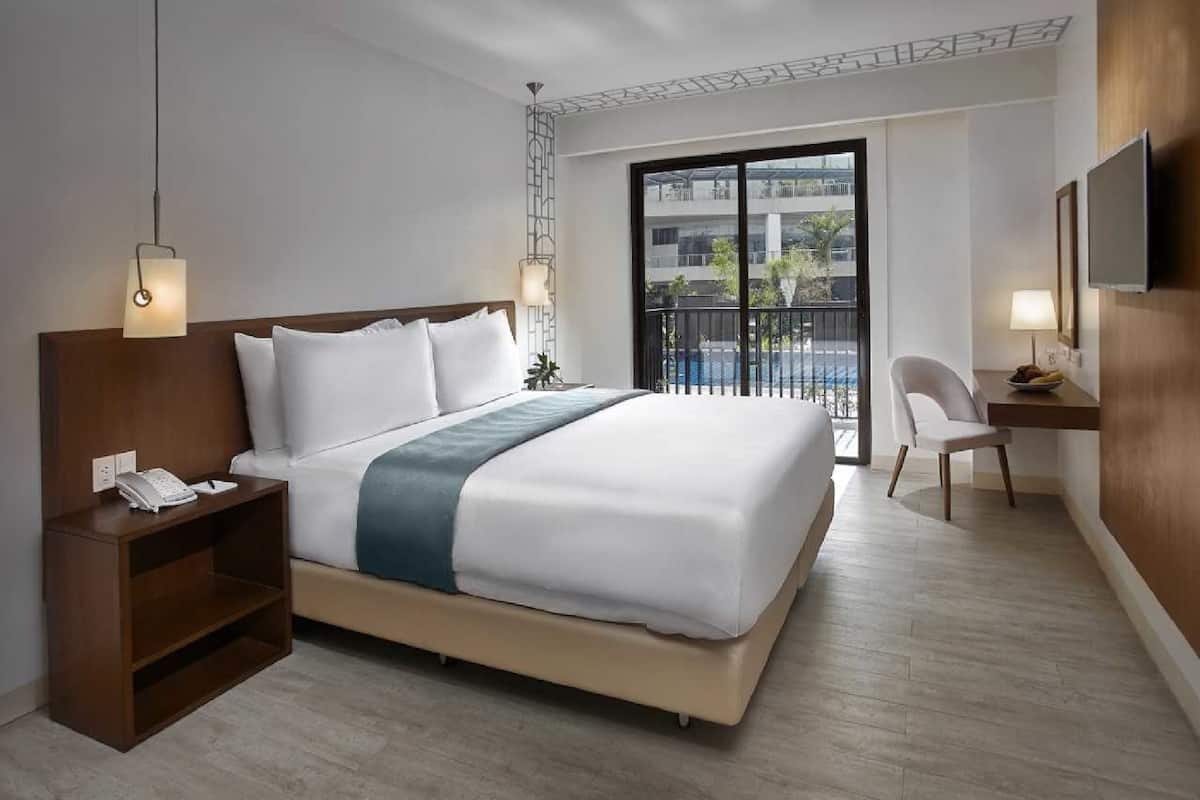 Quarto de hotel com cama grande, mesas de cabeceira, escrivaninha, cadeira e varanda com vista.