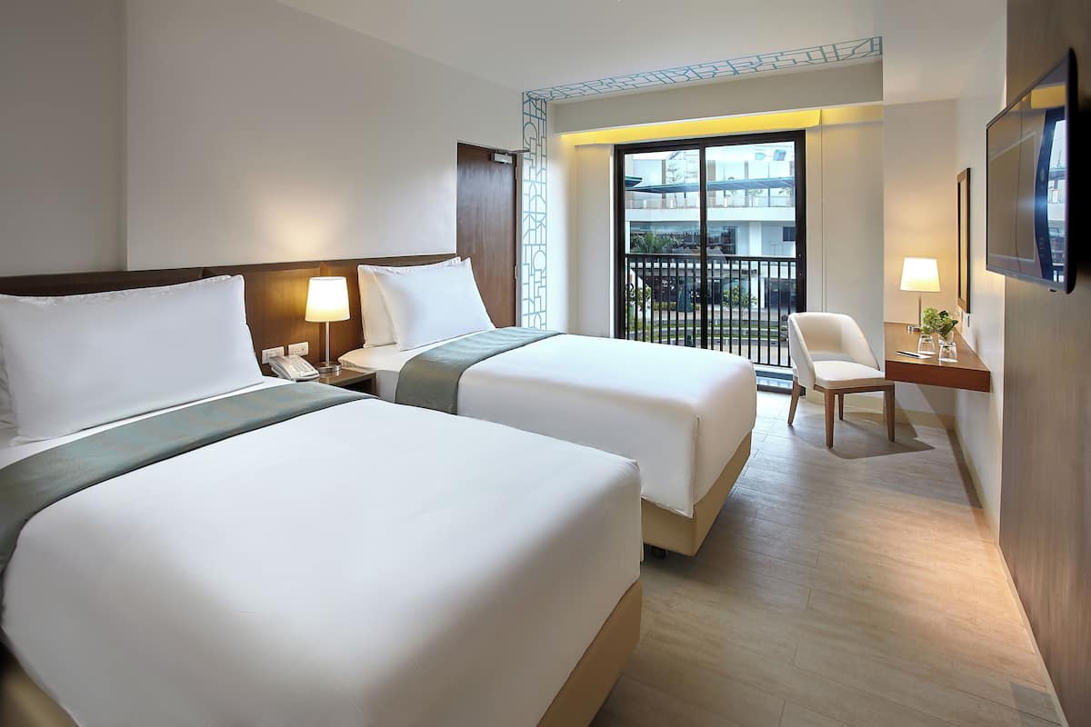 Quarto de hotel com duas camas, uma escrivaninha com cadeira, televisão e varanda com vista para edifícios.