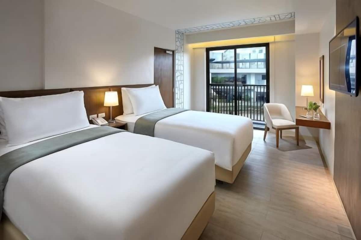 Quarto de hotel com duas camas, uma escrivaninha, uma cadeira, uma televisão e varanda com vista.