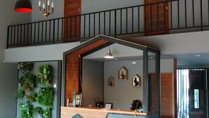 Um hall amplo com balcão de recepção, teto alto com vigas aparentes, um mezanino e uma parede verde com plantas.