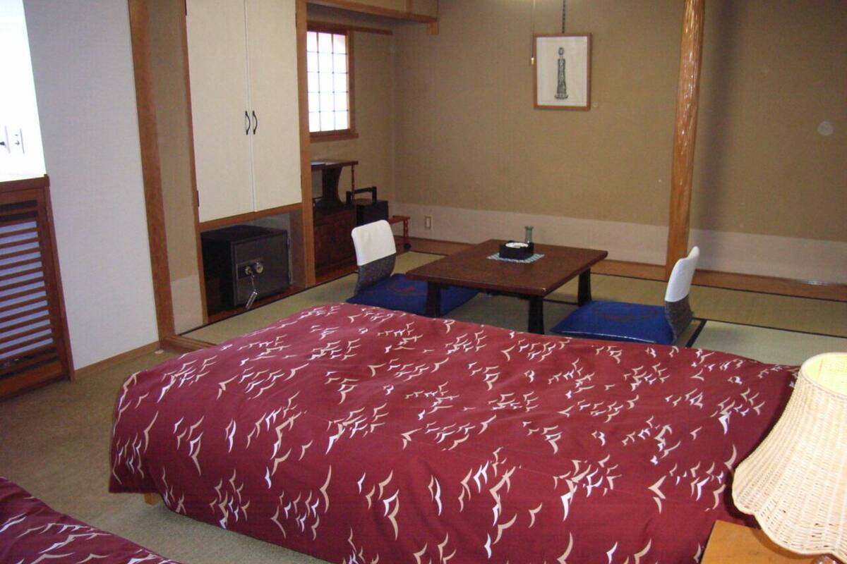Quarto de hotel com uma cama grande, uma escrivaninha, uma cadeira e uma mesinha. Há uma janela com persianas, um quadro na parede e um abajur na escrivaninha.