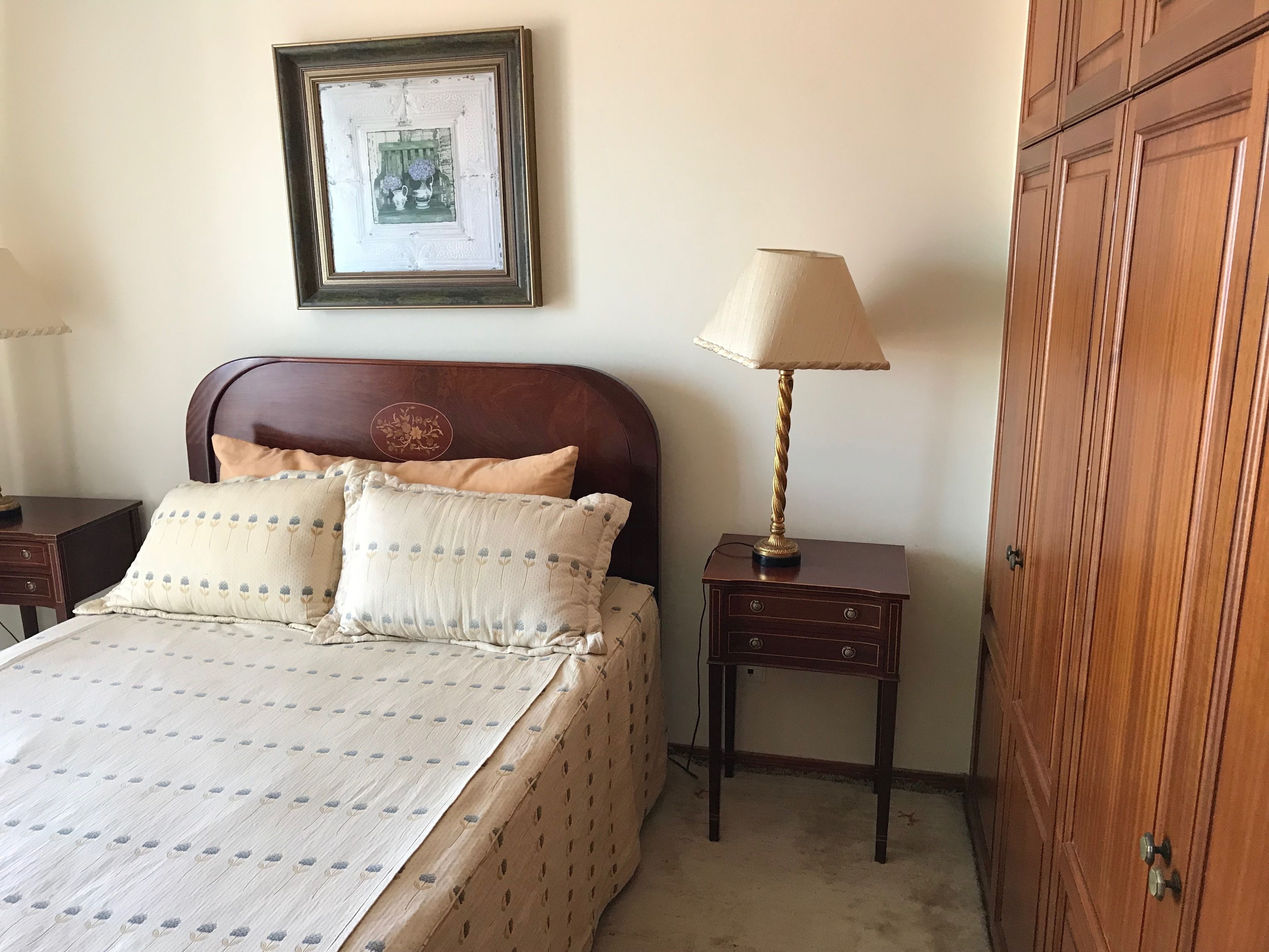 Quarto bem arrumado com cama, mesas de cabeceira, abajur, quadro emoldurado e guarda-roupa de madeira.