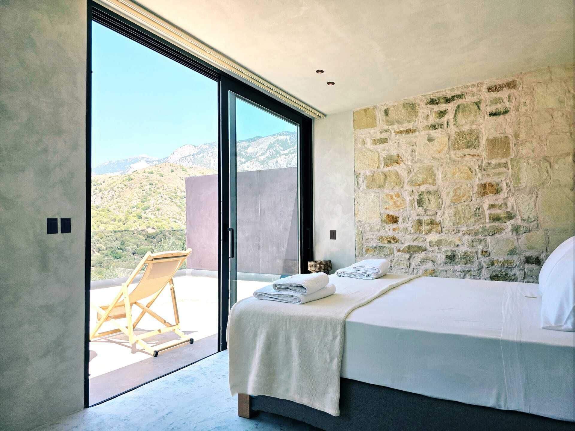 Quarto com cama grande, parede de pedra e vista para uma paisagem montanhosa através de uma porta deslizante.