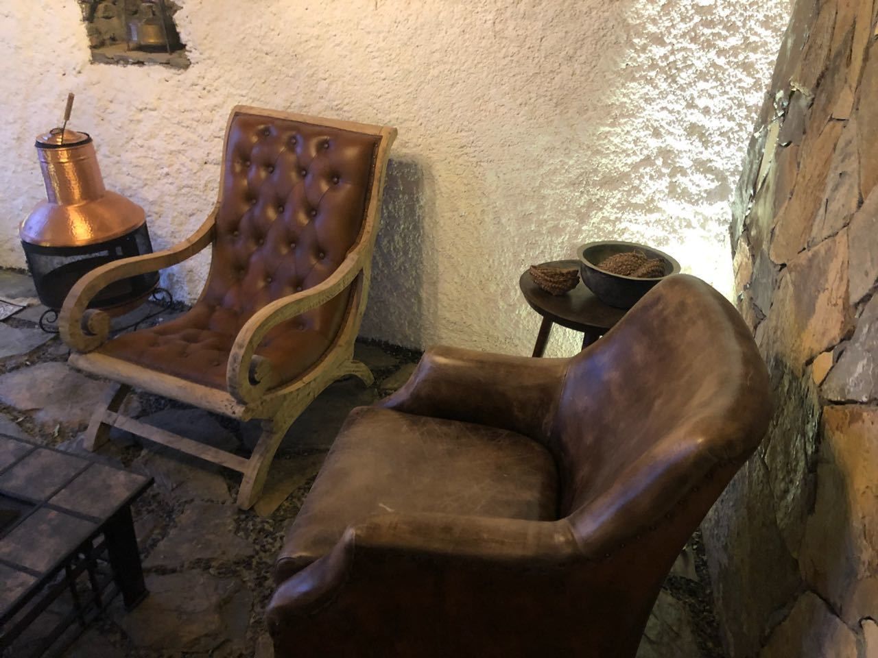 Um quarto rústico com uma poltrona de couro e uma cadeira estofada, uma pequena mesa com uma tigela de metal e uma panela de cobre.
