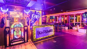 Uma área de bar com um letreiro de neon que diz “HEADLINES”, rodeada por máquinas de fliperama antigas e um espaço de estar com mesas e cadeiras.