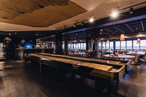 Uma área de bar moderna com um longo balcão de madeira, diversos arranjos de assentos e grandes janelas com vista para o mar.