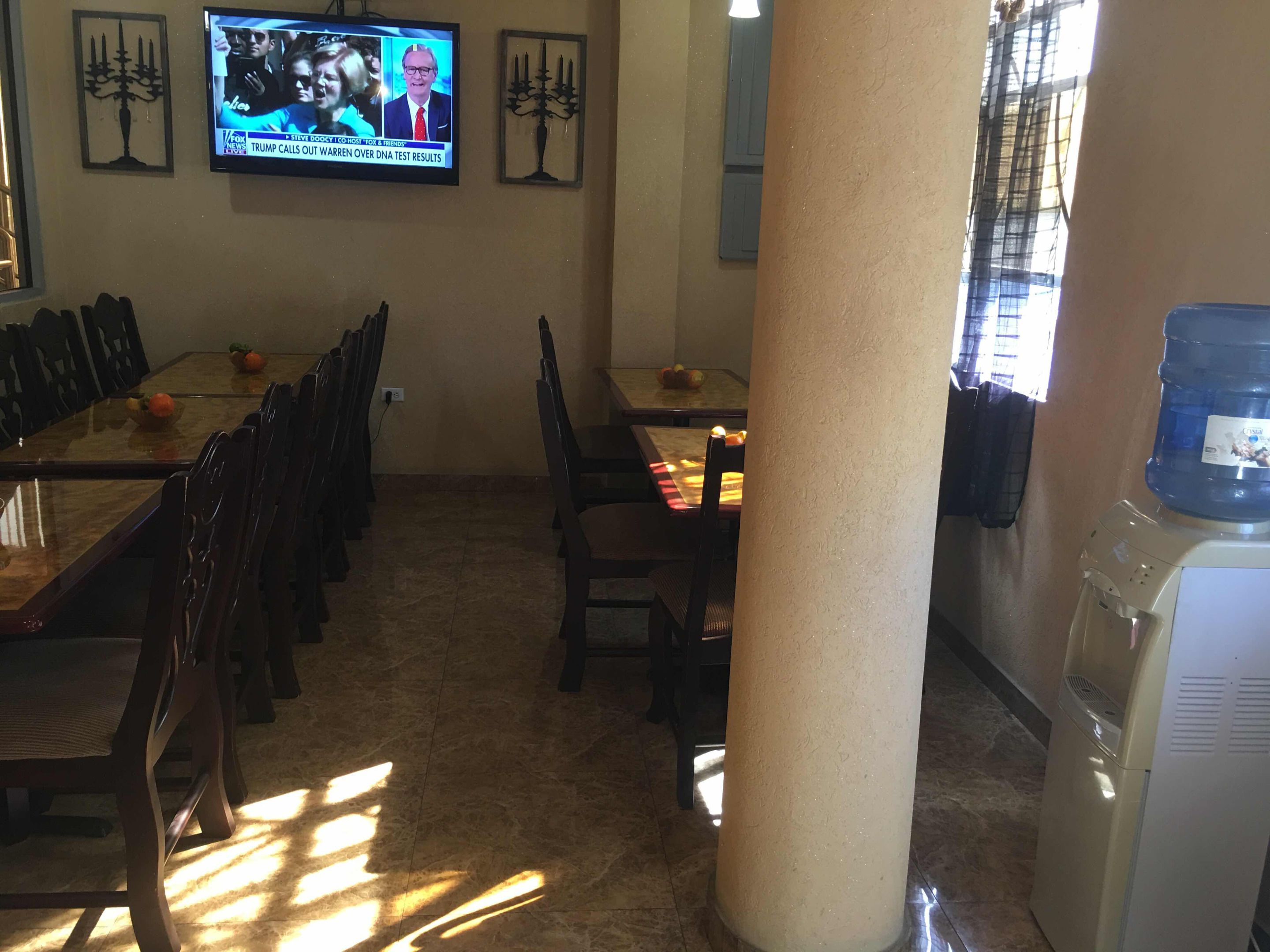 Uma área de refeições com mesas e cadeiras, uma televisão exibindo notícias e um bebedouro.