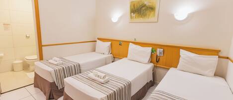 Quarto de hotel com três camas, cabeceira de madeira, um quadro na parede e banheiro com vaso sanitário e pia.