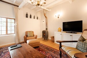 Uma sala de estar acolhedora com um sofá, poltrona, mesa de centro de madeira e uma televisão. Uma lareira é visível ao fundo, e há garrafas em uma prateleira acima dela.