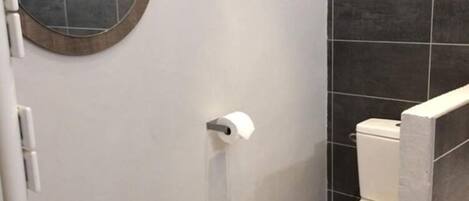 Uma casa de banho com sanita, bidé, suporte para papel higiénico e um espelho redondo na parede.
