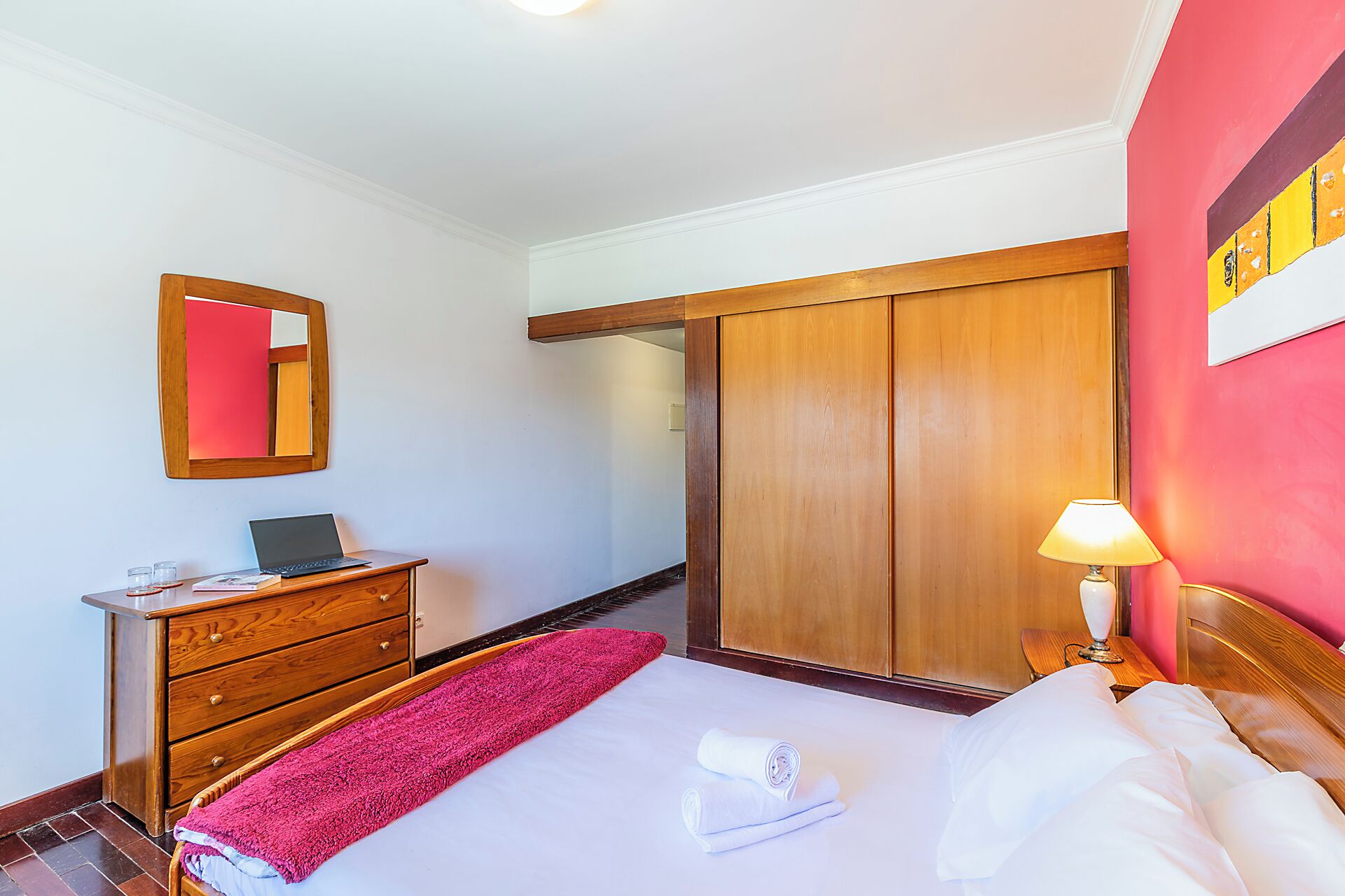 Quarto de hotel com uma cama, um guarda-roupa de madeira, uma escrivaninha com um laptop, um espelho e uma luminária.