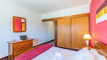 Quarto de hotel com uma cama, um guarda-roupa de madeira, uma escrivaninha com um laptop, um espelho e uma luminária.