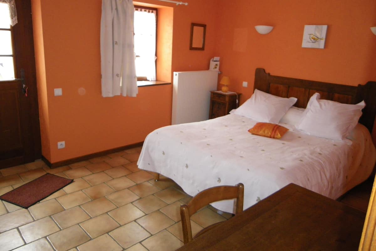 Quarto com cama, cadeira de madeira, mesinha, radiador, janela com cortinas e luminária na parede.