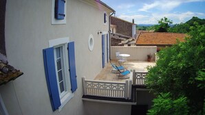 Um edifício de dois andares com uma varanda, um pequeno pátio com mesa e cadeiras, e vista para a paisagem circundante.