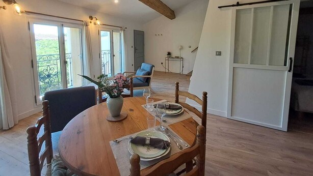 Uma área de refeições com mesa de madeira posta para dois, um vaso com flores e vista para a varanda através das portas deslizantes abertas.