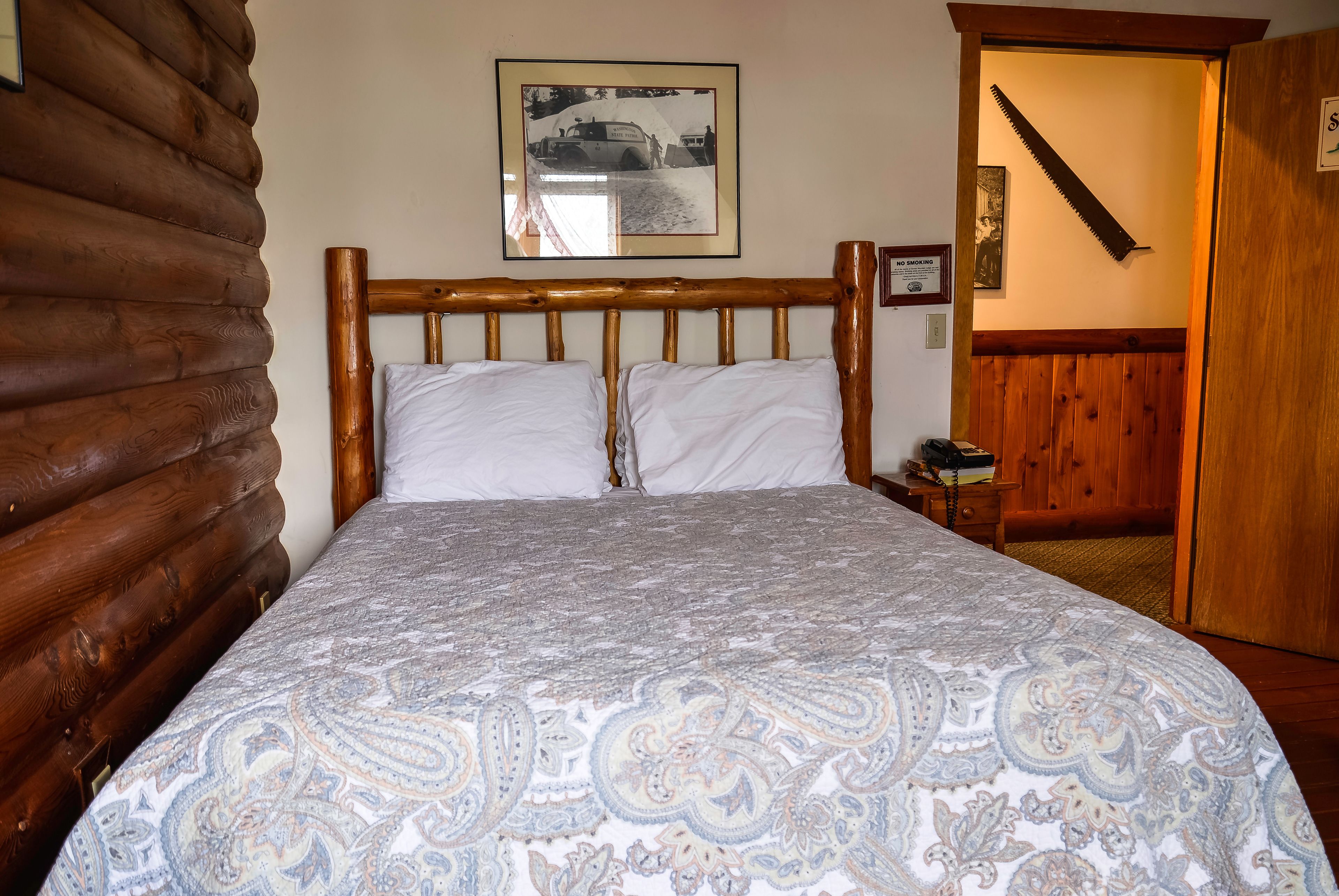 Uma cama de madeira com colcha estampada, um quadro na parede e uma porta de madeira com um serrote pendurado.