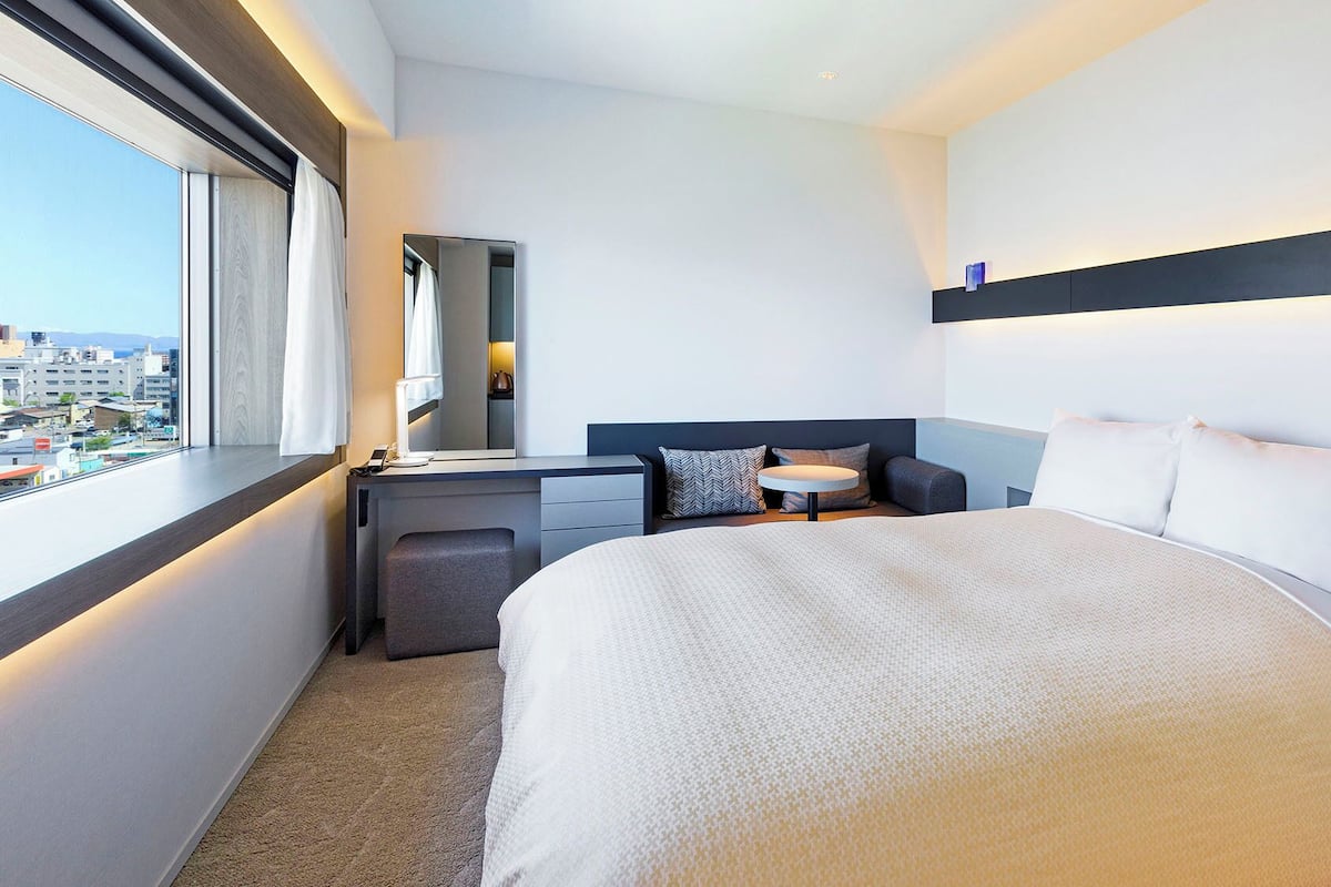 Quarto de hotel com uma cama grande, uma escrivaninha, uma cadeira, uma mesa redonda, um espelho e vista para a cidade.