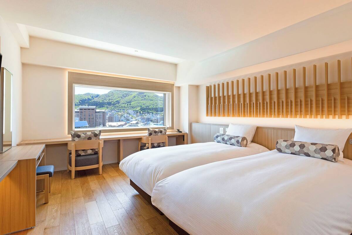 Quarto de hotel com duas camas, uma secretária e uma janela grande com vista para a cidade.