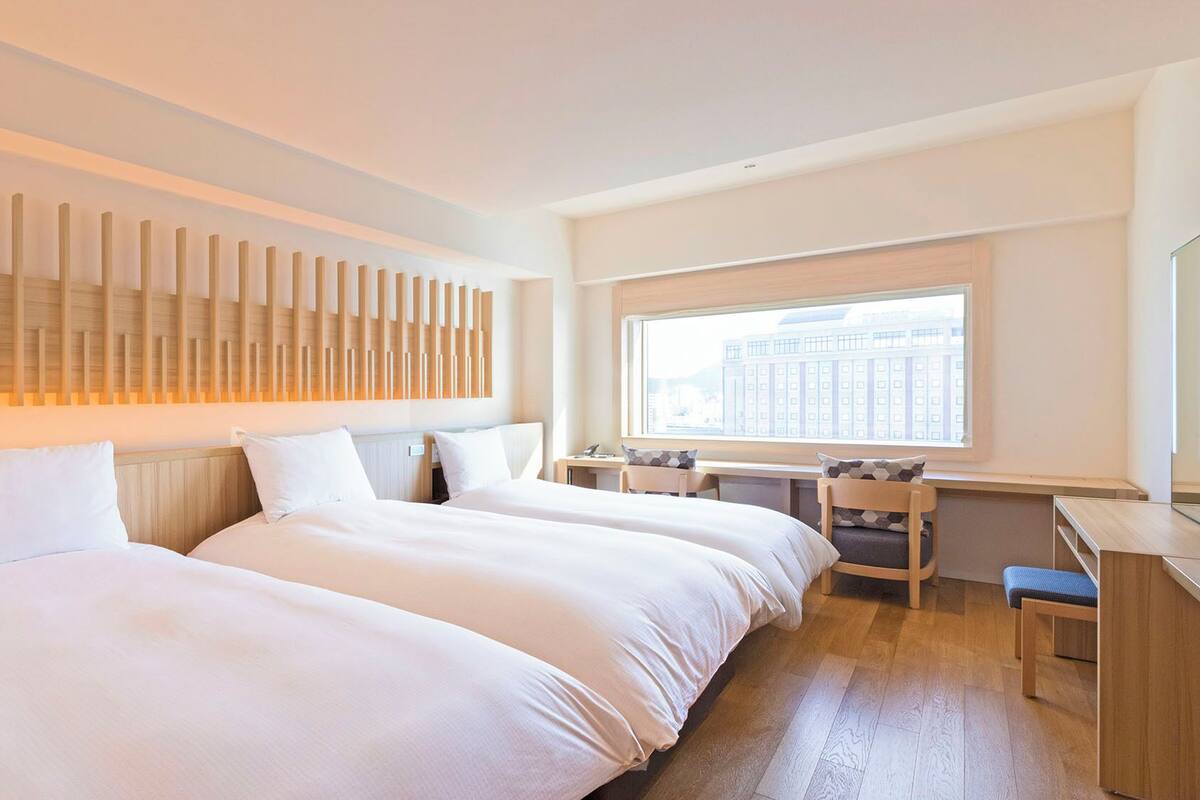Quarto de hotel com duas camas, uma escrivaninha e uma janela com vista para a cidade.