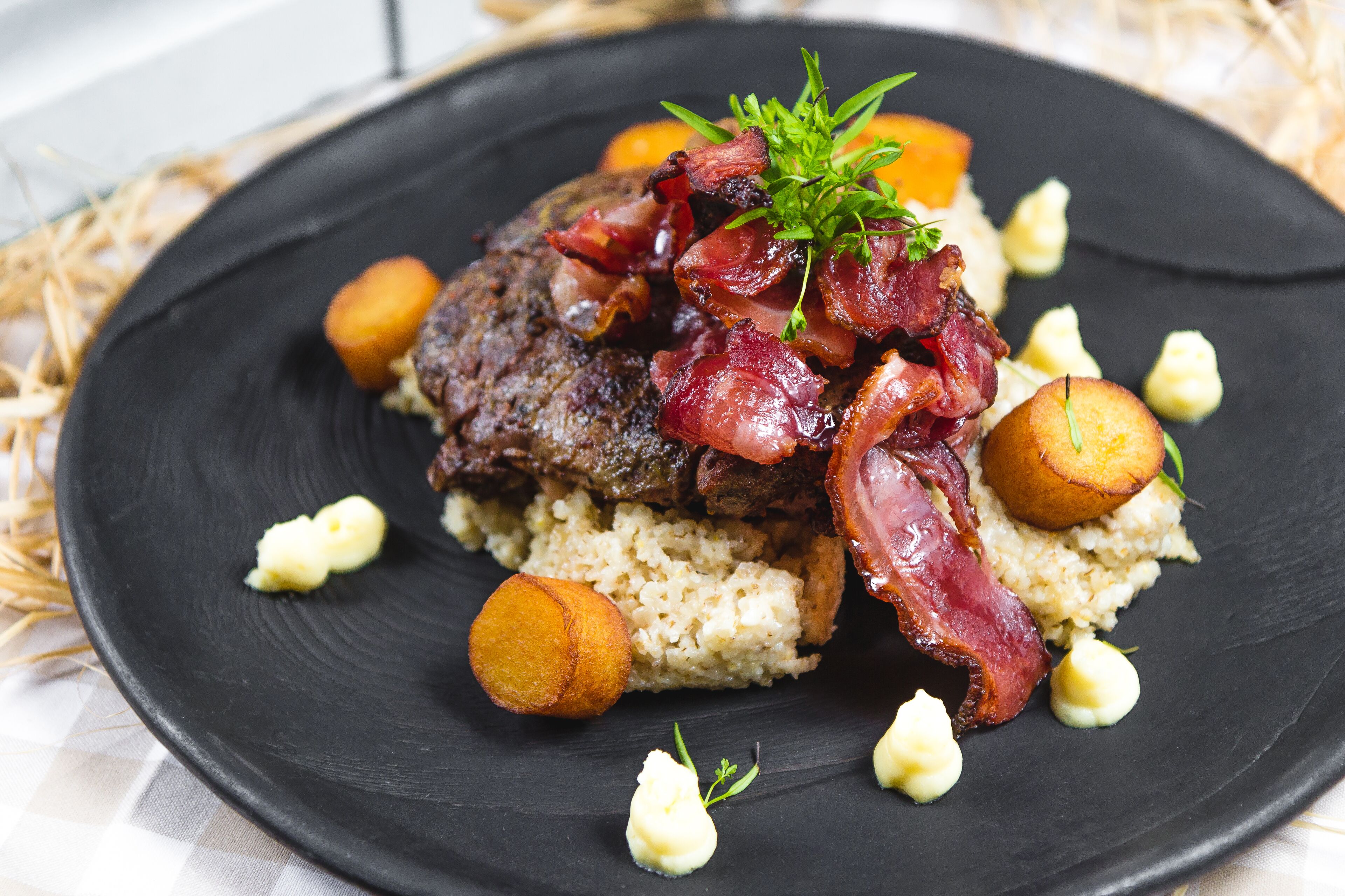 Um prato negro com bife coberto de bacon, acompanhado por batatas assadas e um enfeite.