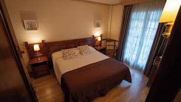 Quarto de hotel com uma cama grande, duas mesas de cabeceira com abajures, cabeceira de madeira e uma cadeira perto da janela.