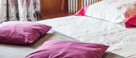 Quarto com cama, duas almofadas, uma cadeira, um radiador e uma cortina.
