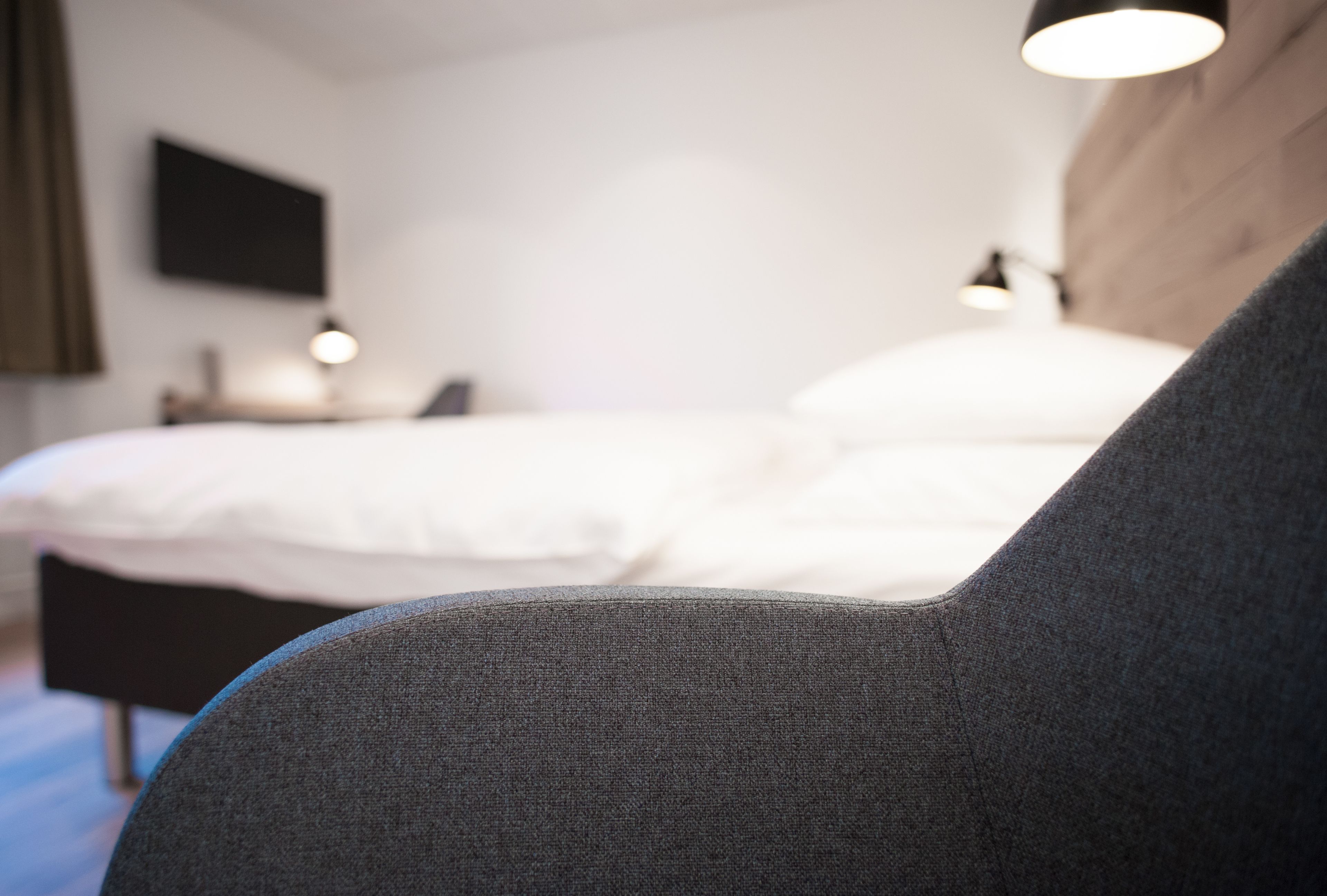 Quarto de hotel moderno com uma cama bem arrumada, uma poltrona cinza escura e uma mesa de cabeceira com abajur.