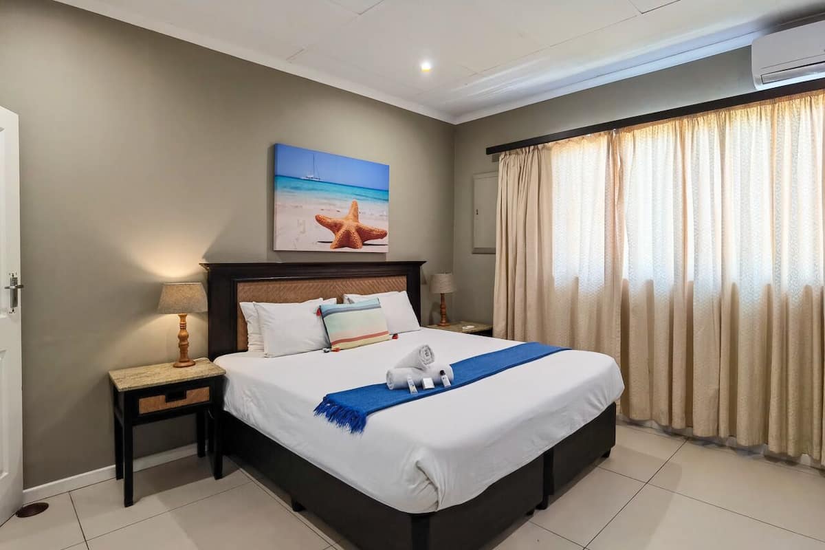 Quarto de hotel com cama, mesa de cabeceira, luminária, quadro de uma praia e janela com cortinas.
