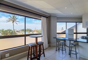 Cozinha moderna com uma janela ampla, área de refeições com bancos e vista para palmeiras e edifícios.