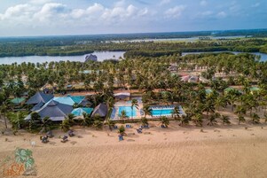 Vista aérea de um resort com vários edifícios, uma piscina grande e uma área de praia com palmeiras.