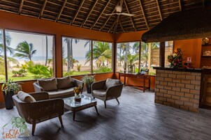 Quarto com telhado de colmo, grandes janelas com vista para uma praia, mobiliário de vime e lareira de tijolos.