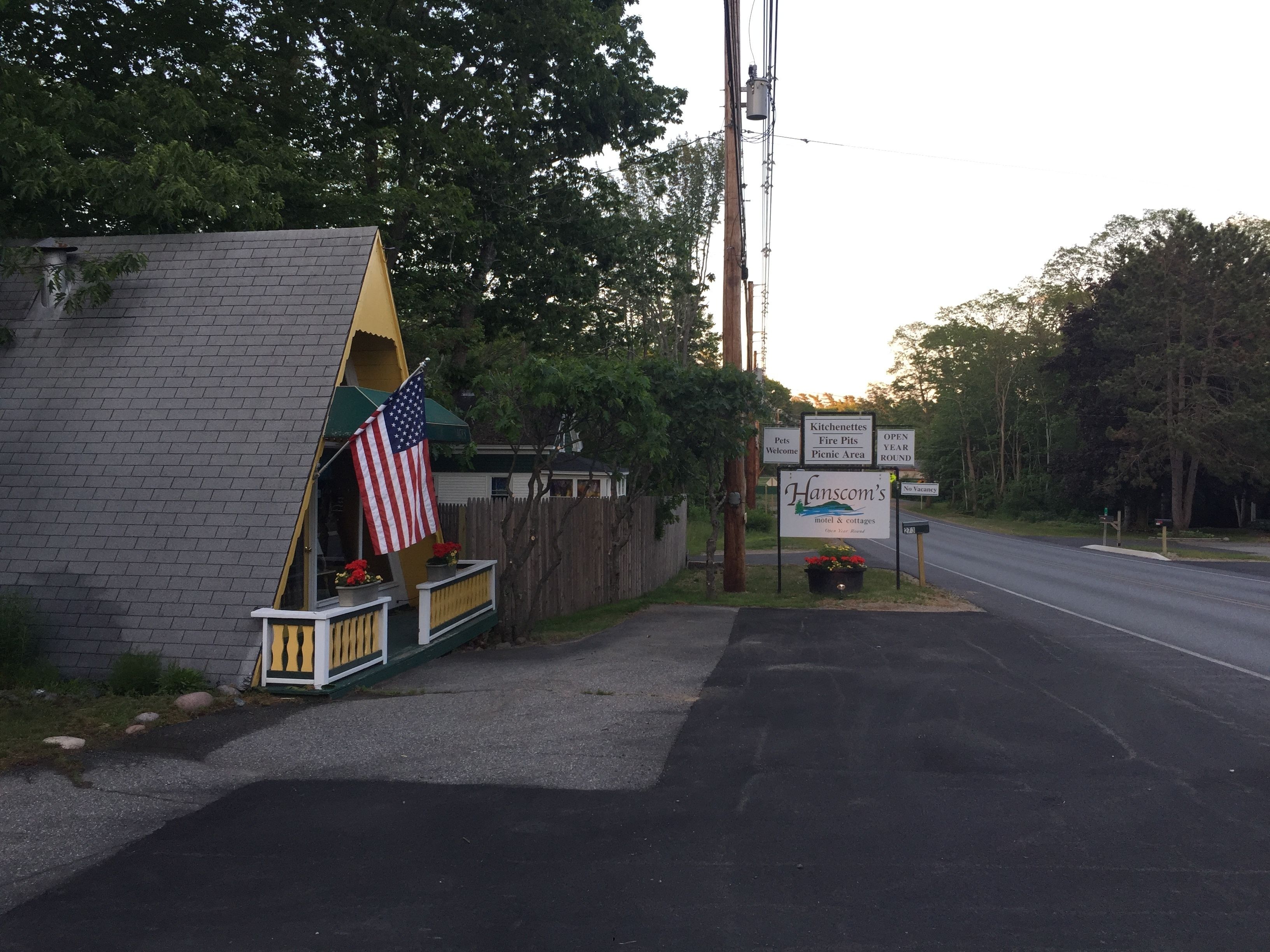 Um motel à beira da estrada com uma pequena exposição de bandeiras americanas, placas com 'Hanscom's Motel & Cottages' e 'Animais de estimação bem-vindos'.