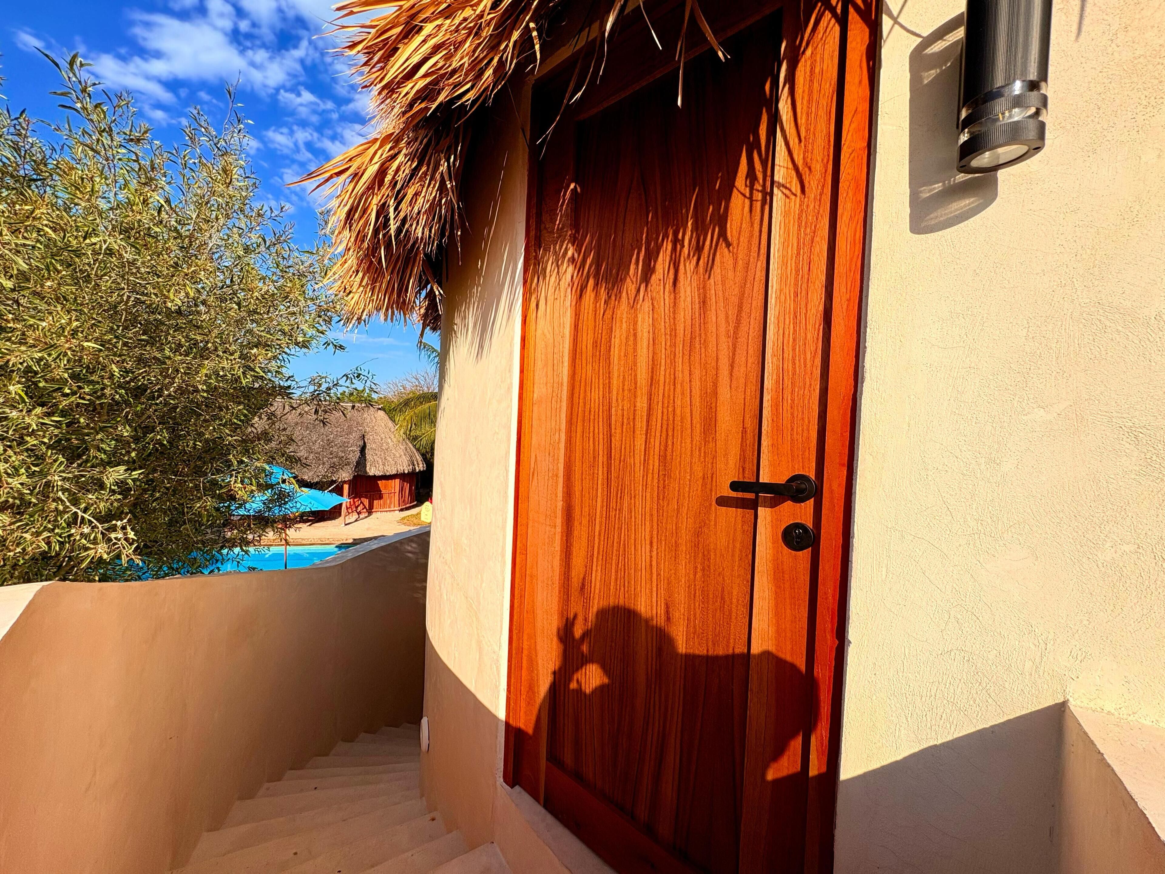 Uma porta de madeira com uma fechadura, uma escadaria que leva a uma área de piscina e uma construção com telhado de palha ao fundo.