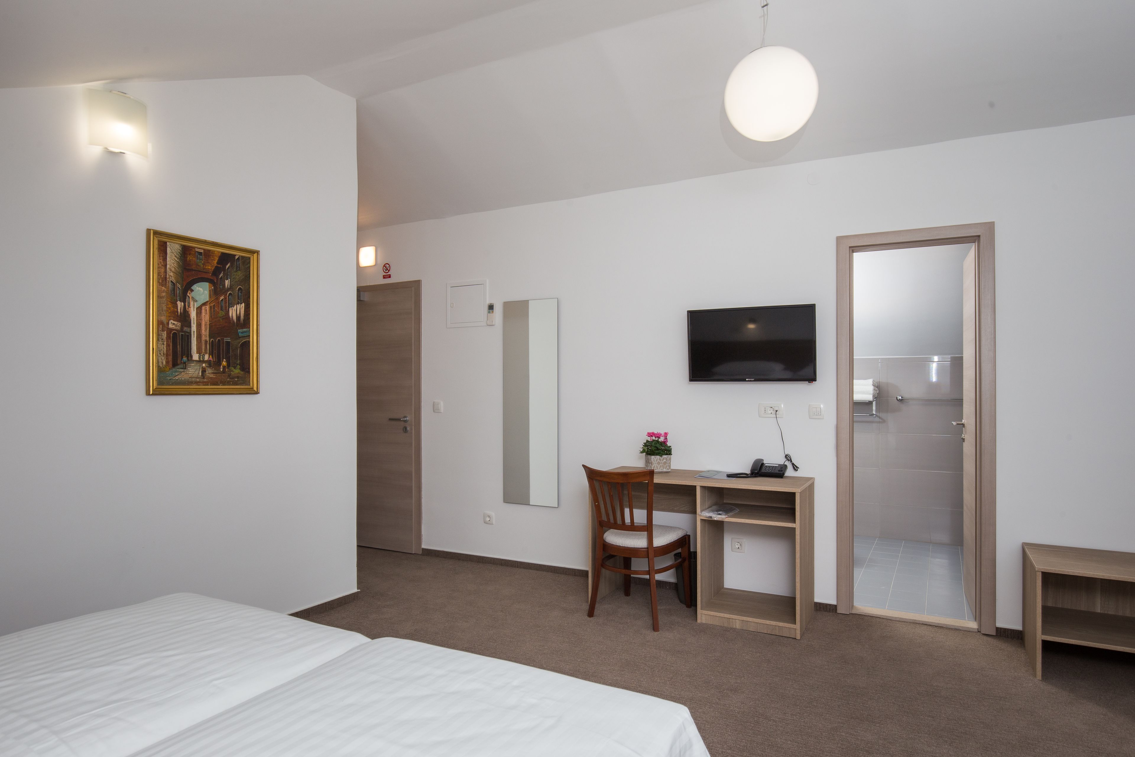 Quarto de hotel com cama, secretária com cadeira, televisão, espelho e porta para uma casa de banho.