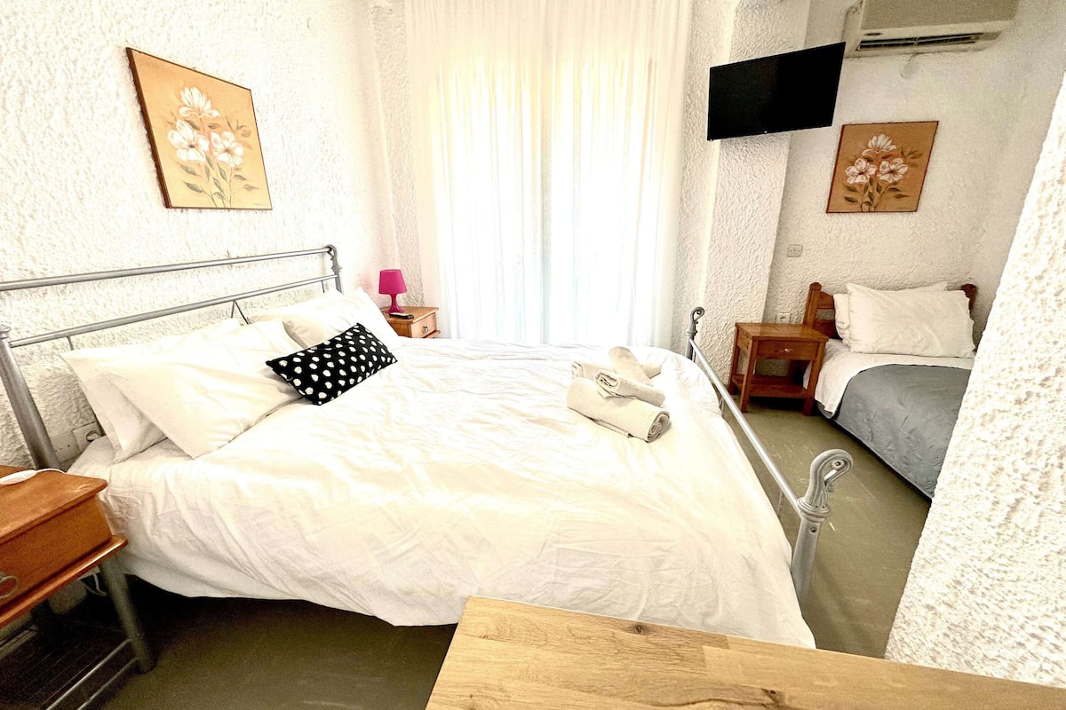 Quarto de hotel com cama, mesa de cabeceira de madeira, escrivaninha, cadeira, televisão fixada na parede e janela com cortinas.