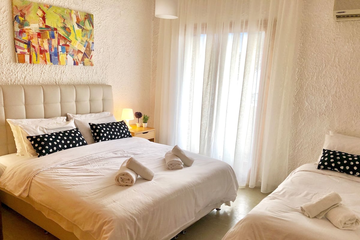 Quarto de hotel com uma cama grande, duas camas menores, roupa de cama branca, mesa de cabeceira com abajur, planta em vaso e um quadro abstrato colorido na parede.