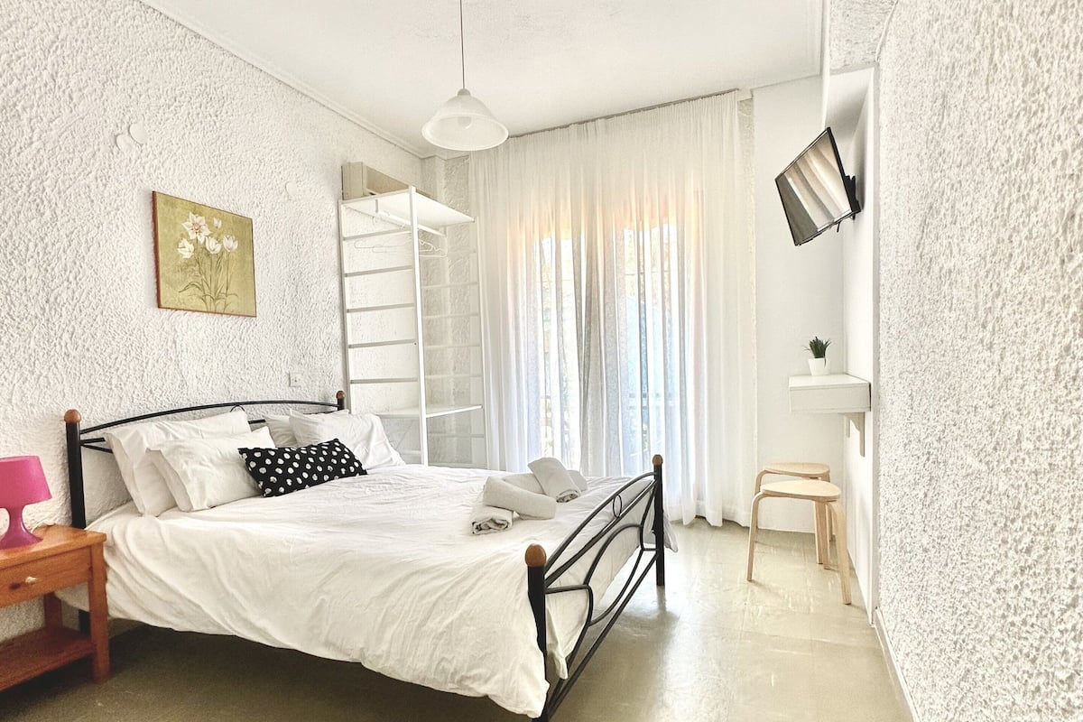 Quarto com cama, mesa de cabeceira, cadeira, escada, luminária pendente e janela com cortinas.