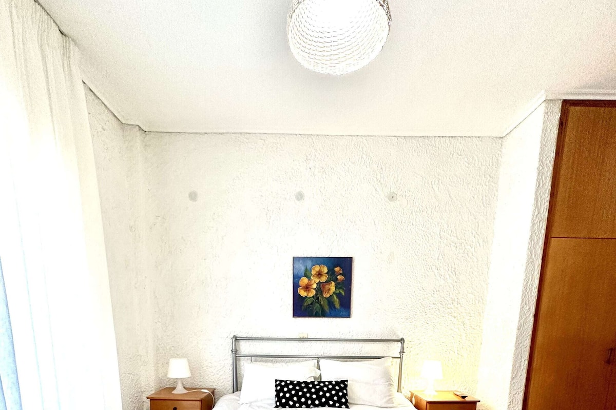 Quarto com estrutura de cama em metal, roupa de cama branca, uma pequena mesa de cabeceira com abajur, um quadro na parede e uma porta de madeira.