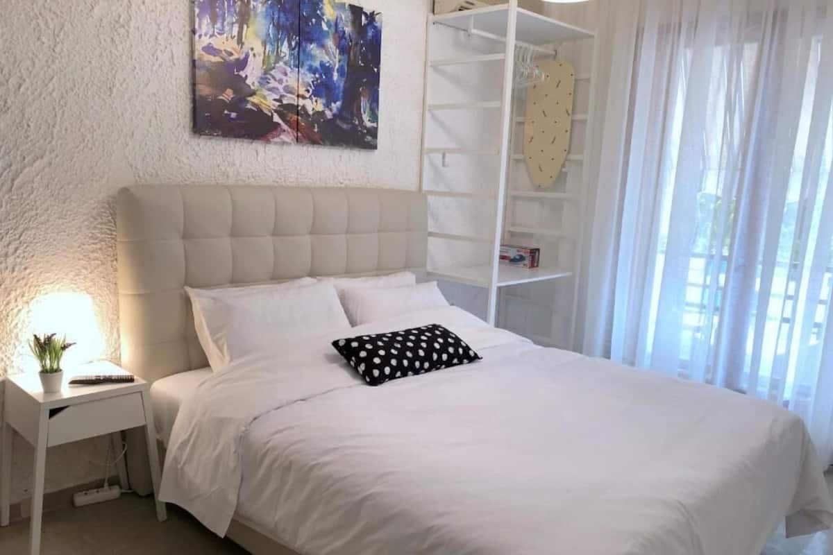 Cama bem arrumada com roupa de cama branca e um travesseiro preto com bolinhas, mesa de cabeceira com abajur e uma pequena planta, e uma estante branca na parede.