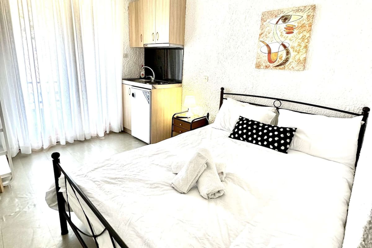 Cama bem arrumada com travesseiro e toalha, uma pequena mesa de cabeceira com abajur, uma cozinha compacta com micro-ondas e armários, e uma janela com cortinas.