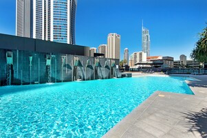 Uma piscina com água azul cristalina, rodeada por edifícios modernos e um horizonte urbano ao fundo.