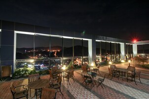 Um pátio externo com mesas e cadeiras, com vista para o horizonte urbano à noite.
