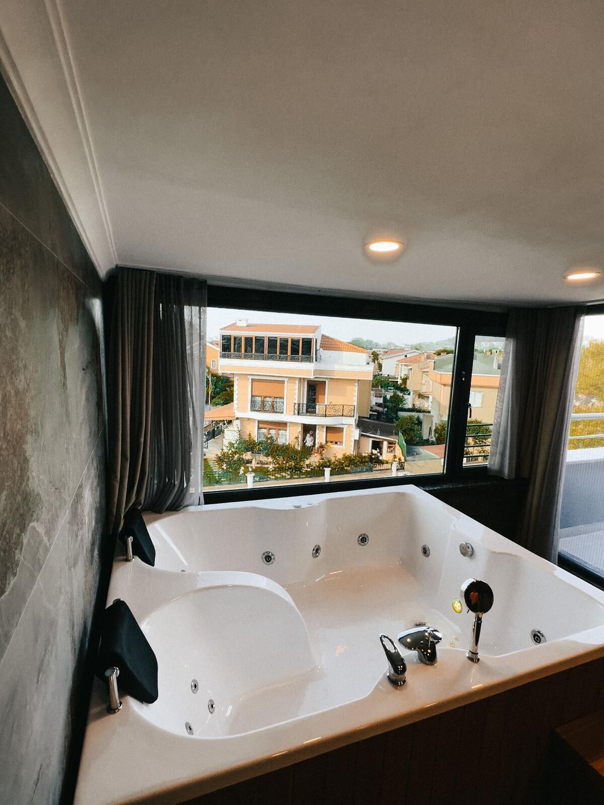 Uma casa de banho moderna com uma banheira grande, uma janela com cortinas e vista para uma área residencial.