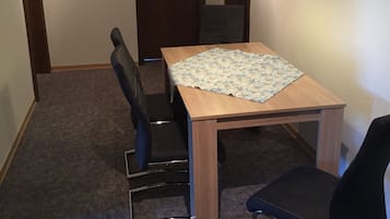 Um quarto pequeno e simples, com uma mesa de madeira, duas cadeiras pretas, piso coberto por carpete e uma porta.
