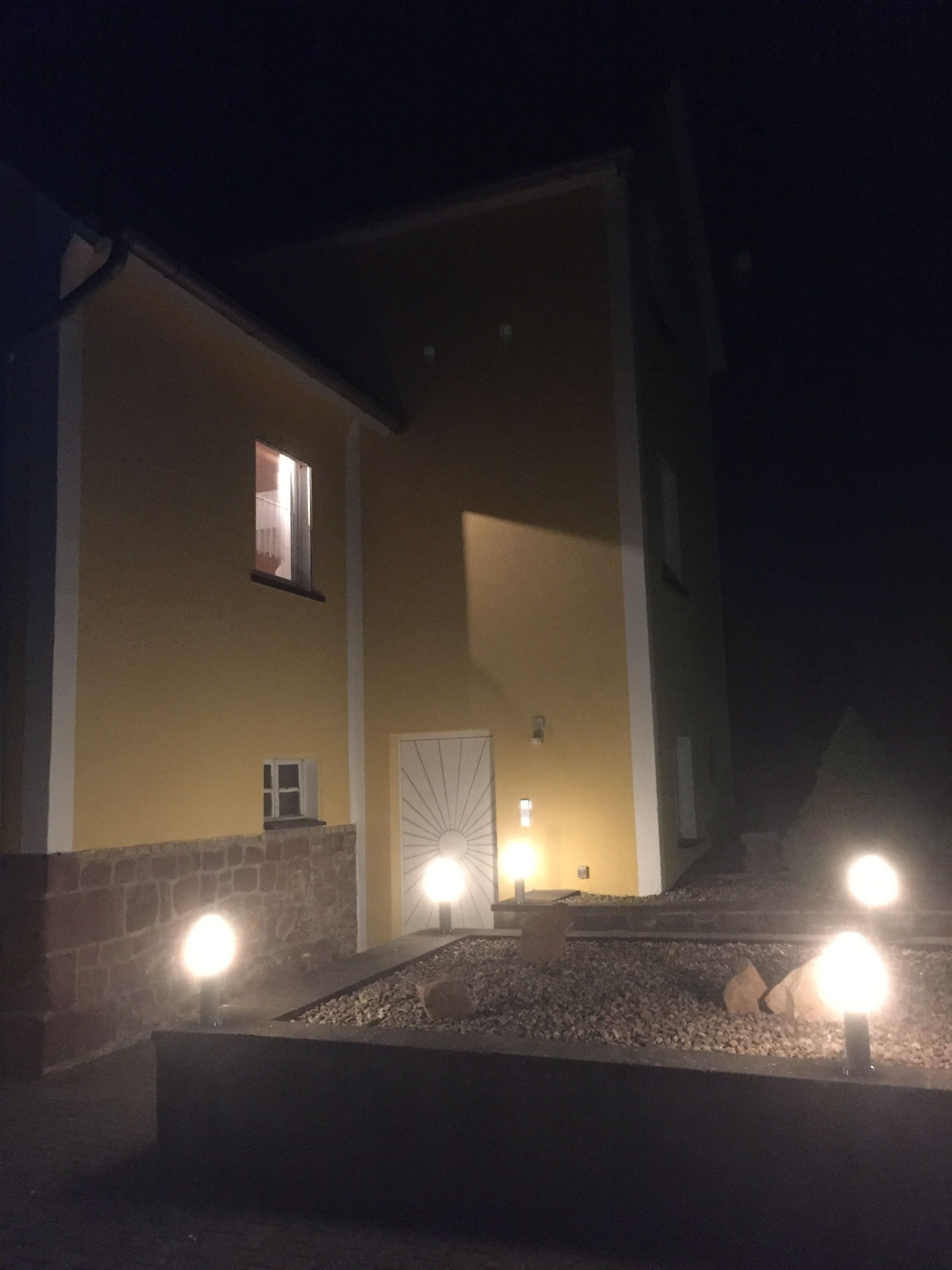Um edifício com fachada amarela, base de pedra e uma porta com design em forma de sol. Três luminárias em primeiro plano.