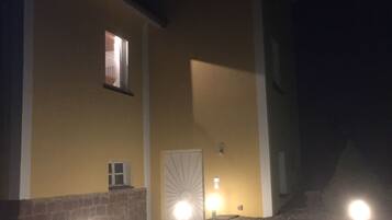 Um edifício com fachada amarela, base de pedra e uma porta com design em forma de sol. Três luminárias em primeiro plano.