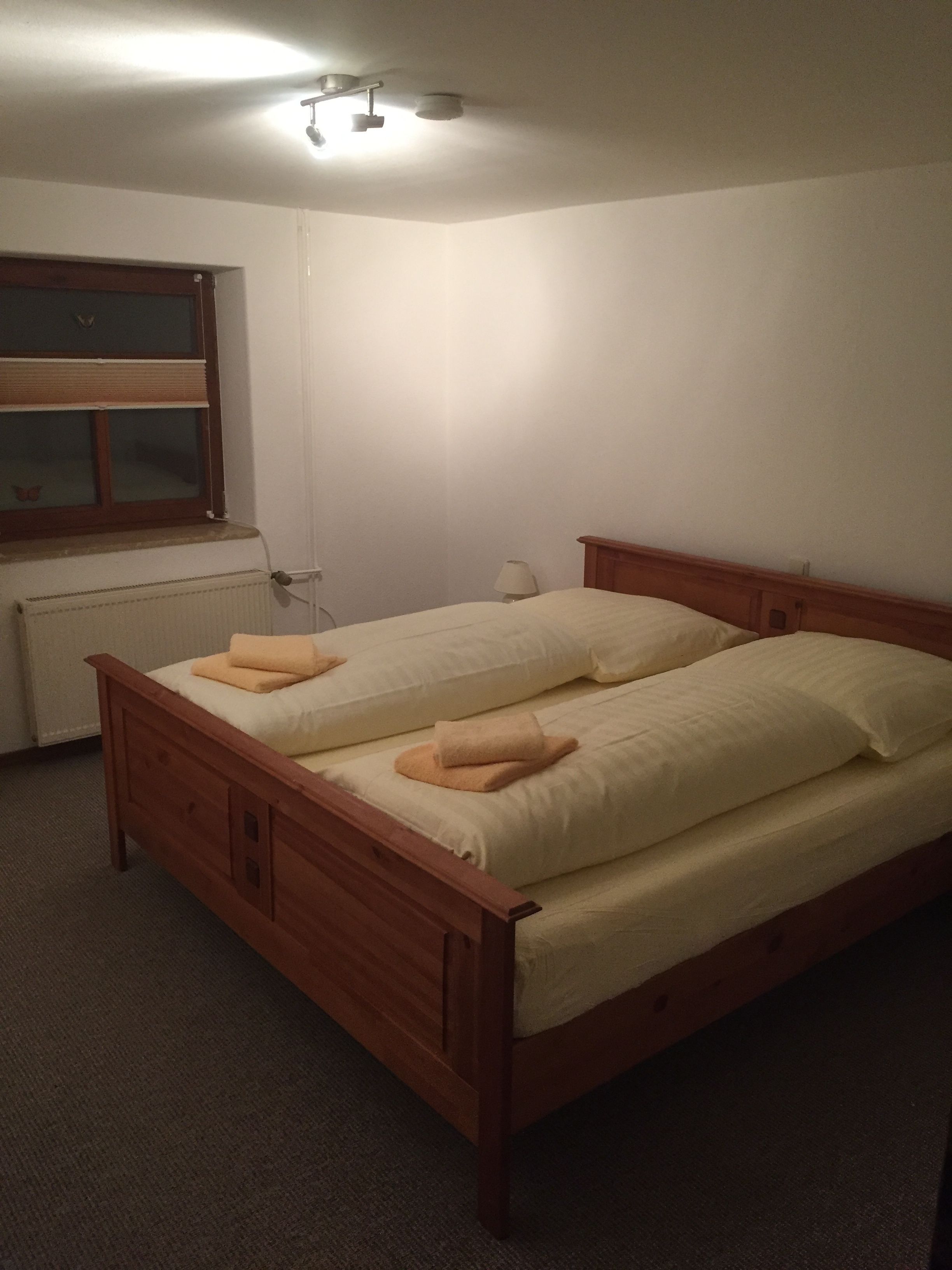 Quarto com cama de madeira, duas almofadas, abajur e janela com persianas.