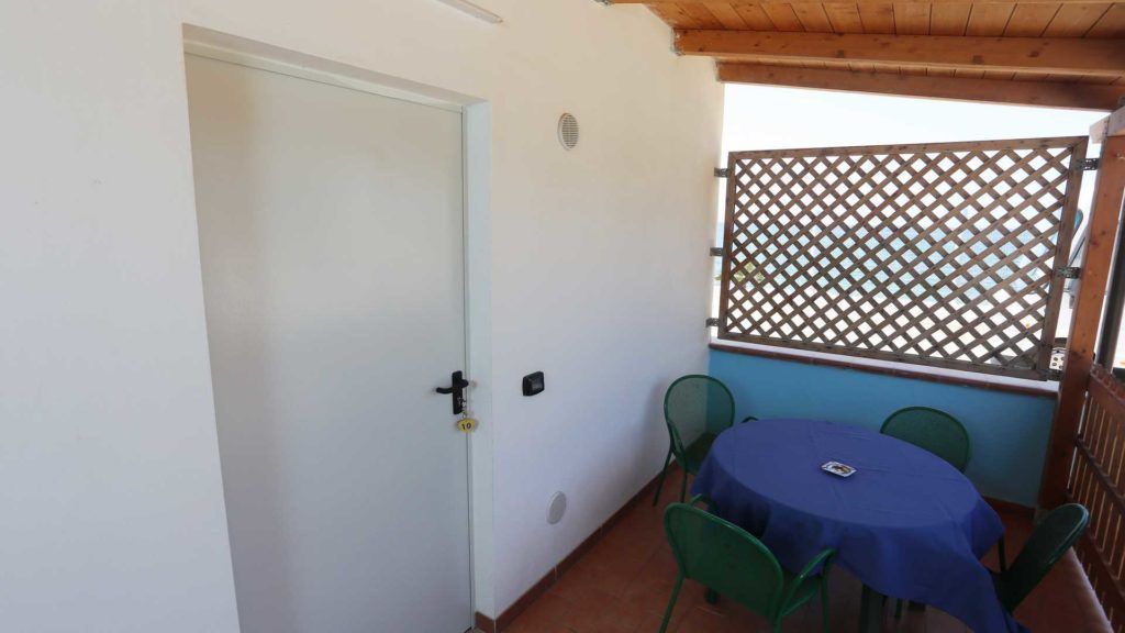 Quarto com porta branca, pequena janela e varanda com mesa azul e cadeiras verdes.