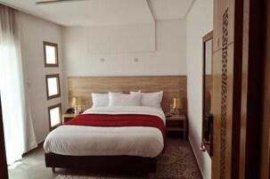 Quarto de hotel com uma cama grande, duas mesas de cabeceira com luminárias, uma cabeceira de madeira e um tapete estampado.