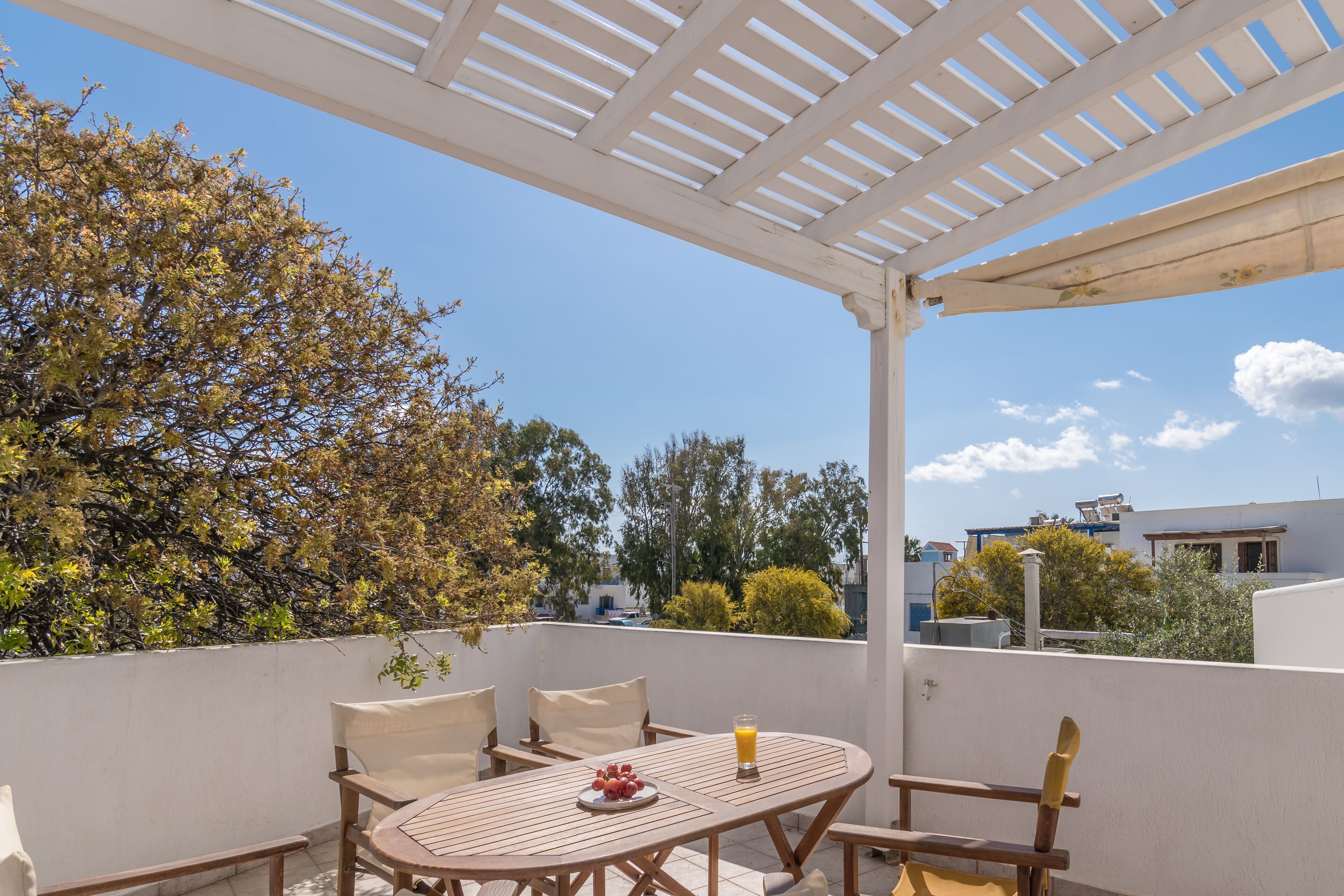 Um pátio externo com mesa e cadeiras de madeira, um copo de suco de laranja e vista para árvores e edifícios ao fundo.